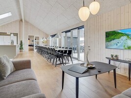 Ampla sala de estar com uma mesa de jantar comprida, móveis modernos e uma grande TV exibindo uma vista panorâmica.