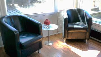 Quarto com duas poltronas pretas, uma mesa redonda pequena com um vaso vermelho e vista para um deck de madeira.