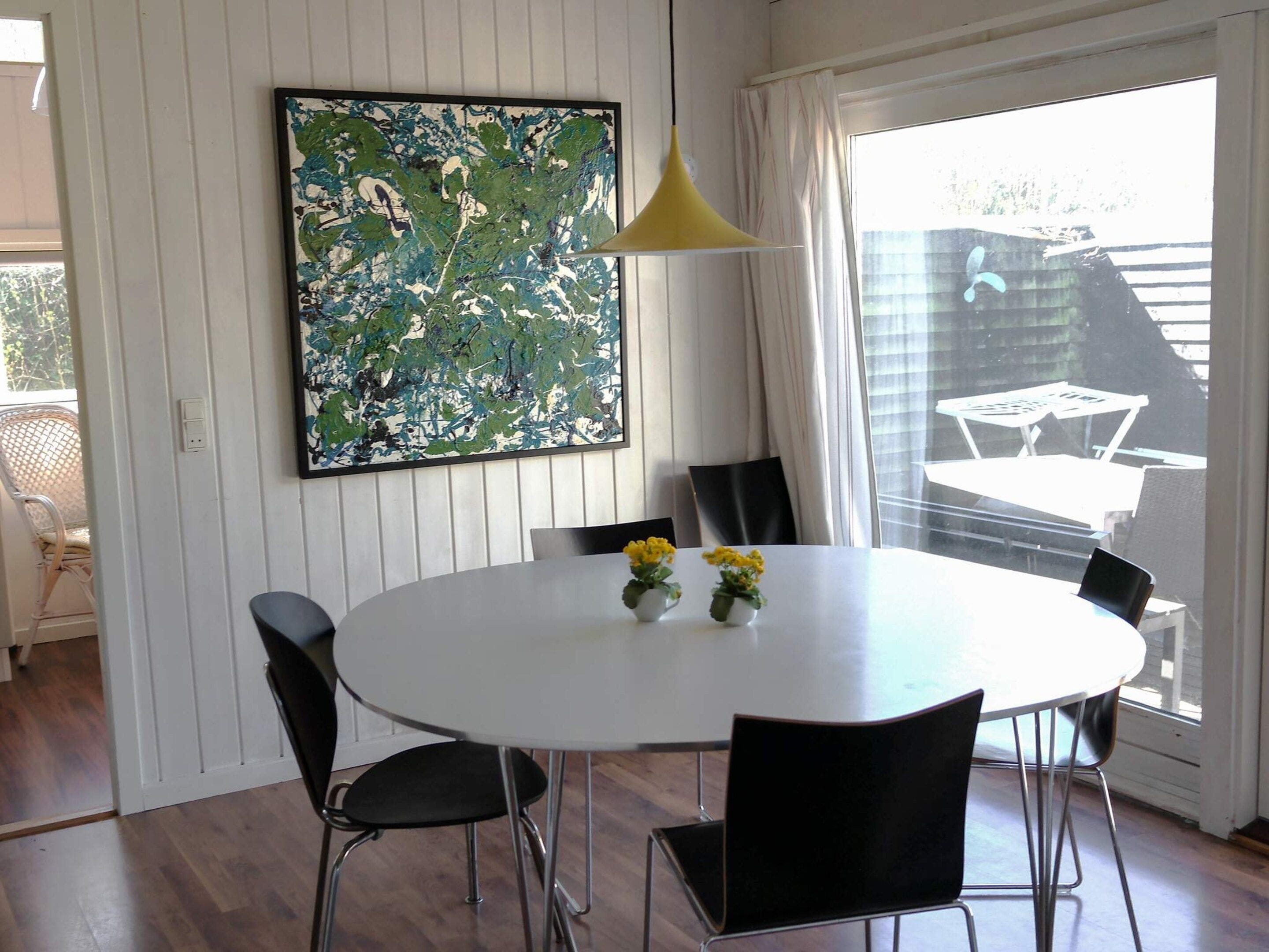 Uma sala de jantar com uma mesa branca, cadeiras pretas, uma luminária pendente amarela e uma grande pintura abstrata na parede.