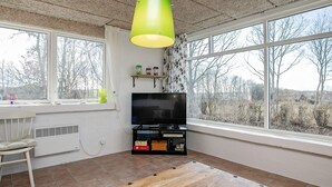 Quarto com televisão, mesa de madeira e amplas janelas com vista para árvores.