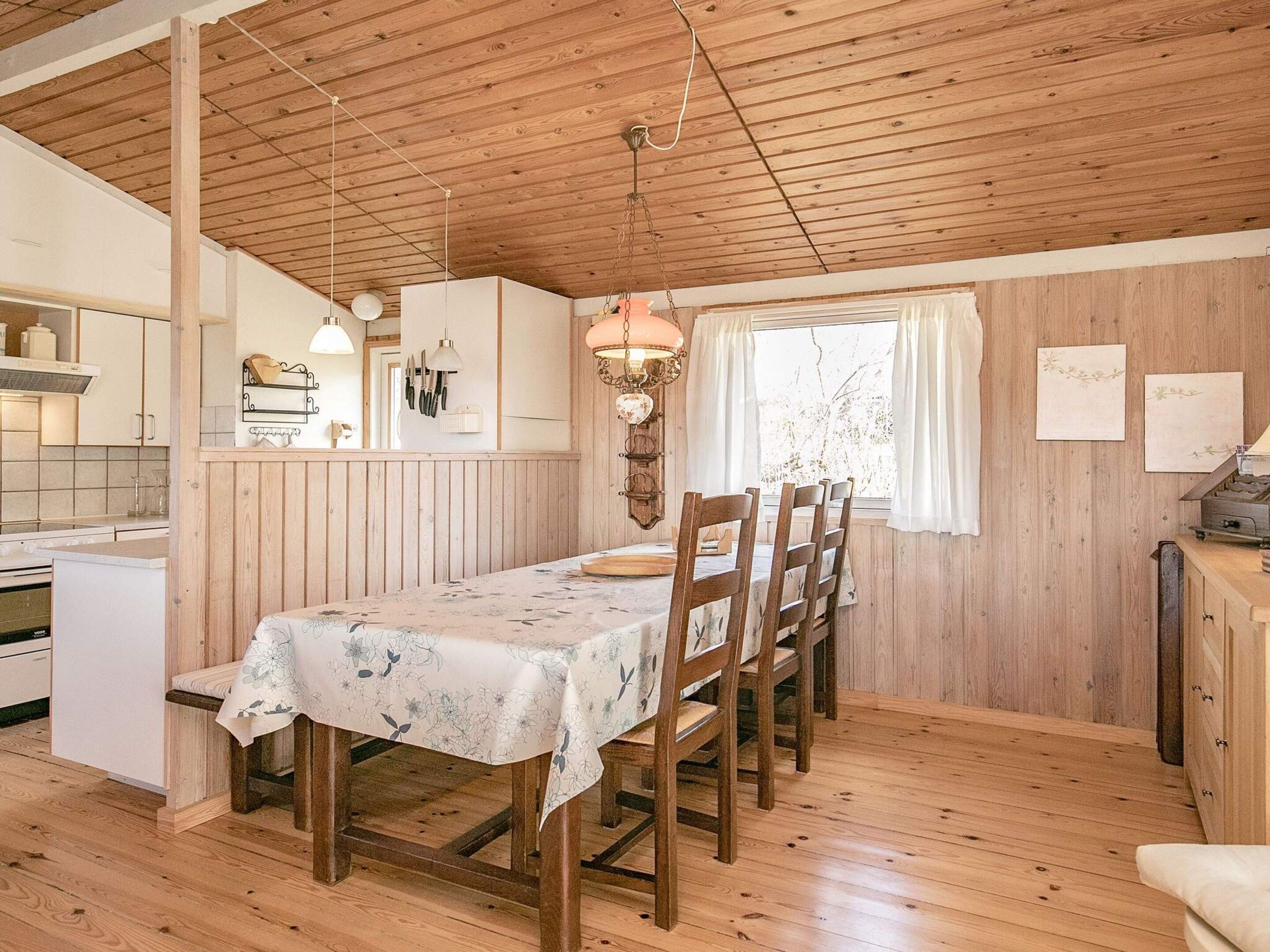 Interior em madeira com uma mesa de jantar posta para quatro, área de cozinha e janela com cortinas.