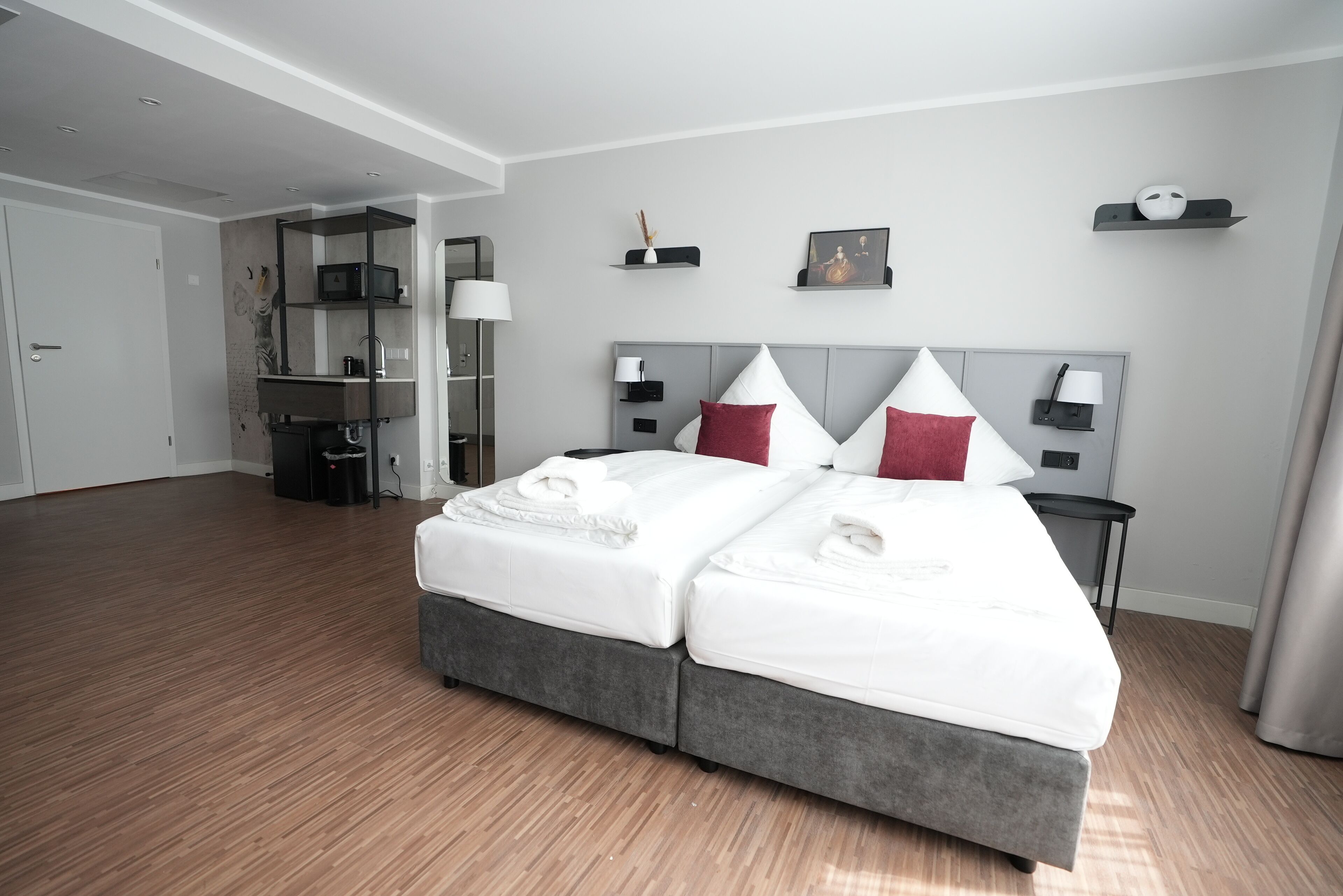 Quarto de hotel moderno com cama de casal, cabeceira cinza, roupa de cama branca e uma pequena mesa de cabeceira com abajur.