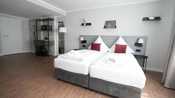 Quarto de hotel moderno com cama de casal, cabeceira cinza, roupa de cama branca e uma pequena mesa de cabeceira com abajur.