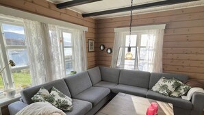 Uma sala de estar aconchegante com um sofá modular cinza, uma mesa de centro de madeira e vista para um lago através de grandes janelas com cortinas brancas.