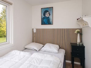Quarto com cama, travesseiros, mesa de cabeceira, vaso com flores, quadro e janela.