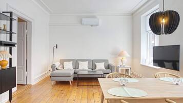 Uma sala de estar moderna com um sofá cinza, uma mesa de jantar de madeira com cadeiras, uma televisão de tela plana e uma luminária pendente.