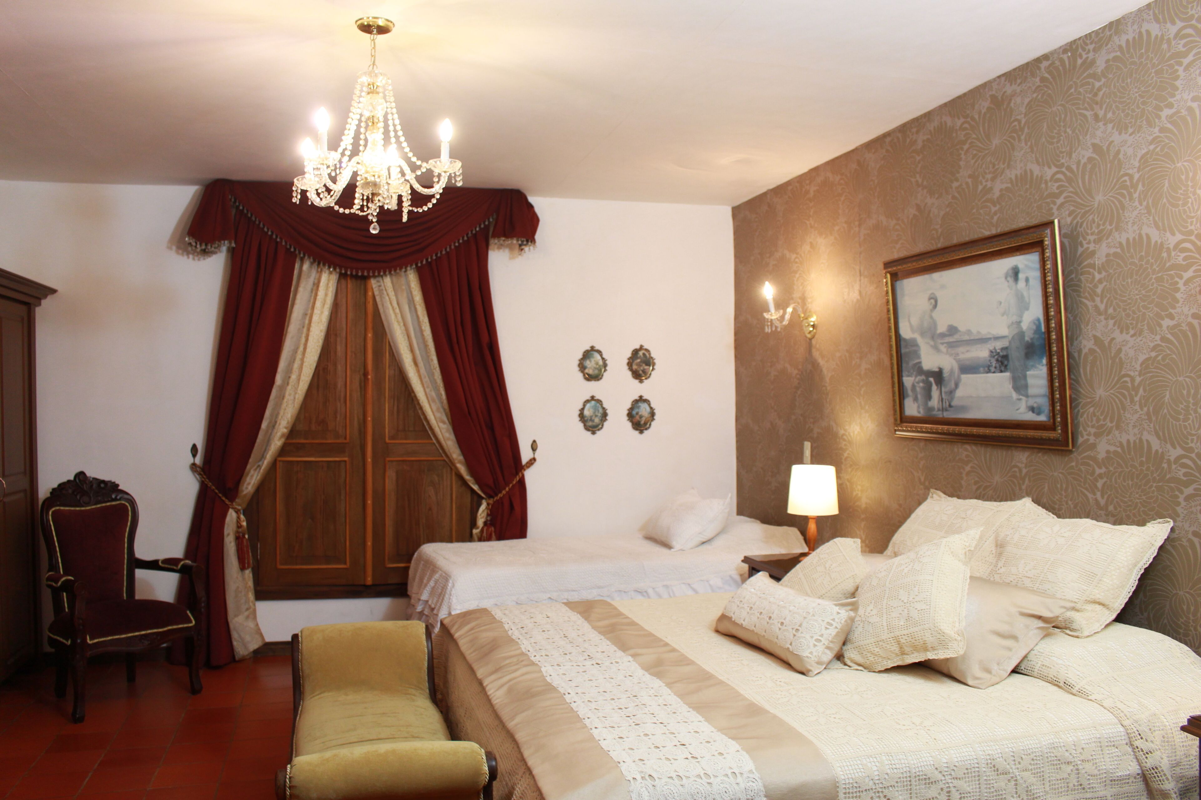 Quarto de hotel com lustre, cama com várias almofadas, uma cadeira, mesa de cabeceira com abajur e um quadro na parede.