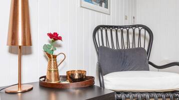 Um quarto com uma mesa de madeira escura, um candeeiro de cobre, um jarro de metal com uma rosa vermelha, uma tigela de metal e uma cadeira de metal preta com almofada branca.