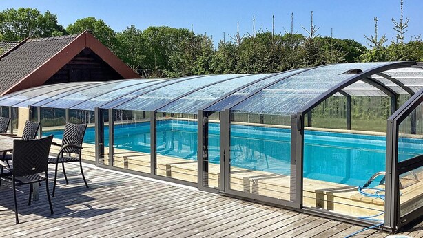 Piscina coberta com deck de madeira, estrutura de vidro e mobiliário para área externa.