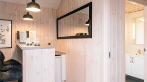 Quarto com paredes em painel de madeira, uma escrivaninha com espelho, uma cadeira e uma porta que dá para outro cômodo.