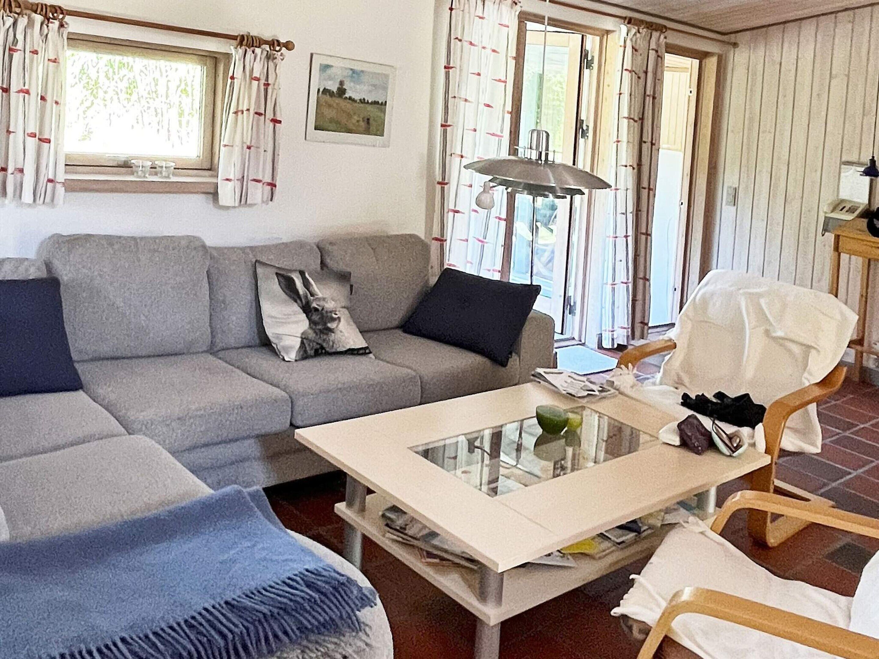 Sala de estar com sofá, mesa de centro e uma cadeira. Há uma janela com cortinas e um quadro na parede.