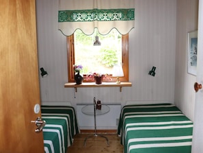 Quarto com duas camas de solteiro, uma janela com cortina de renda verde, uma mesinha com abajur e uma porta.