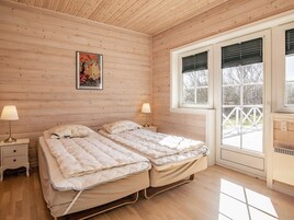 Quarto aconchegante com paredes de madeira e uma janela grande. Há duas camas com roupa de cama branca e duas mesas de cabeceira com luminárias. Um quadro emoldurado está pendurado na parede acima das camas.