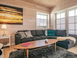 Uma sala de estar aconchegante com um sofá seccional cinza escuro, uma mesa de centro de madeira e um tapete preto e branco. Há duas janelas com persianas, um quadro na parede e um abajur em uma mesa lateral.