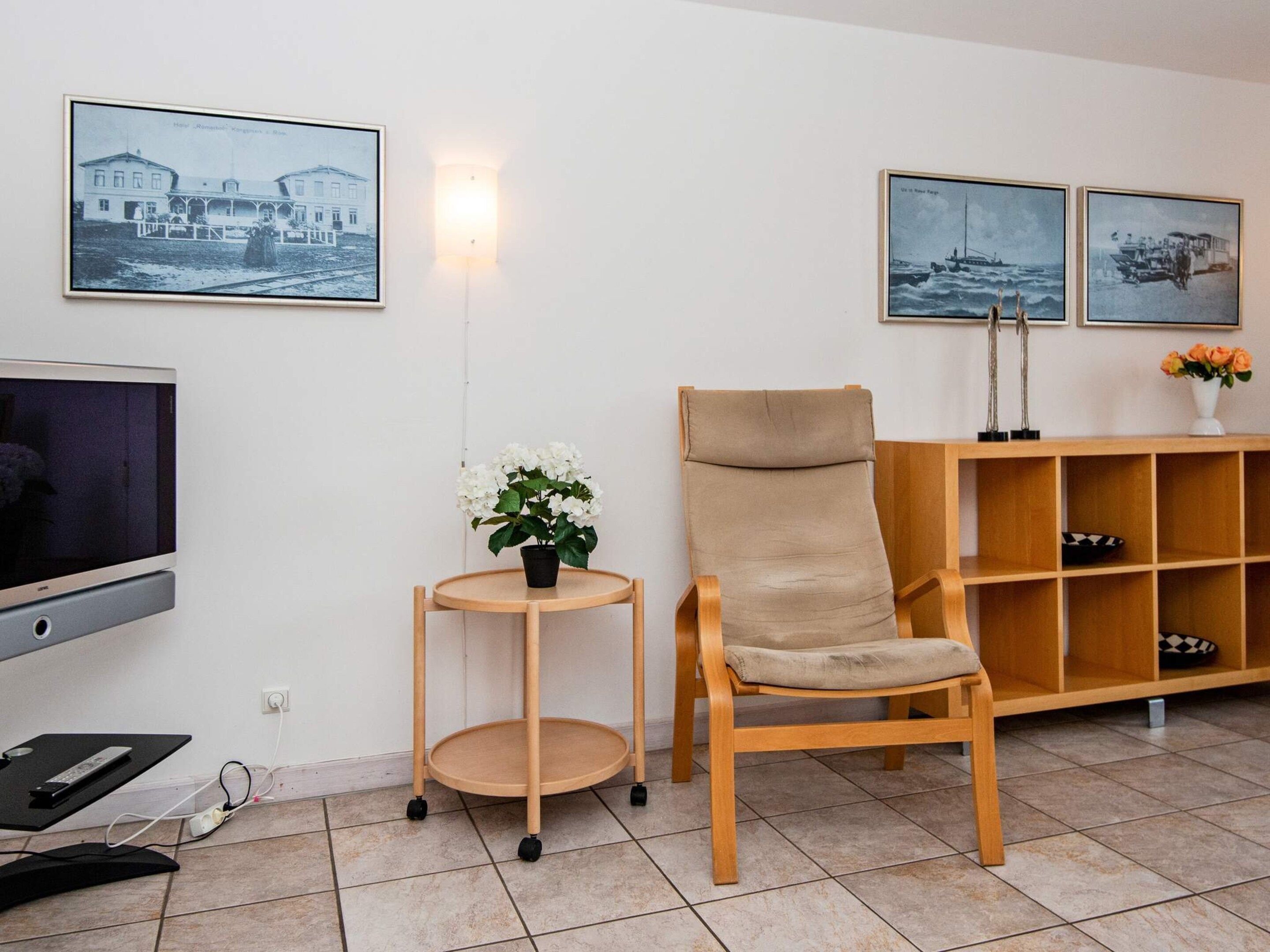 Uma sala de estar com televisão, uma cadeira de madeira, uma pequena mesa com planta em vaso e uma estante de madeira com objetos decorativos.
