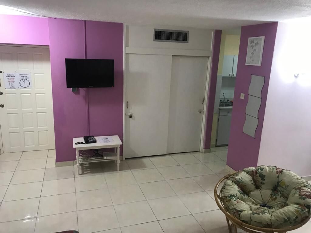 Um quarto com porta fechada, televisão na parede, uma pequena mesa com telefone e papéis, uma cadeira com padrão floral e piso de azulejos.
