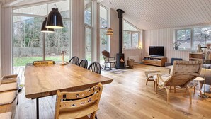 Uma sala de estar espaçosa com uma grande mesa de madeira, várias cadeiras, lareira, televisão e janelas amplas com vista para o exterior.