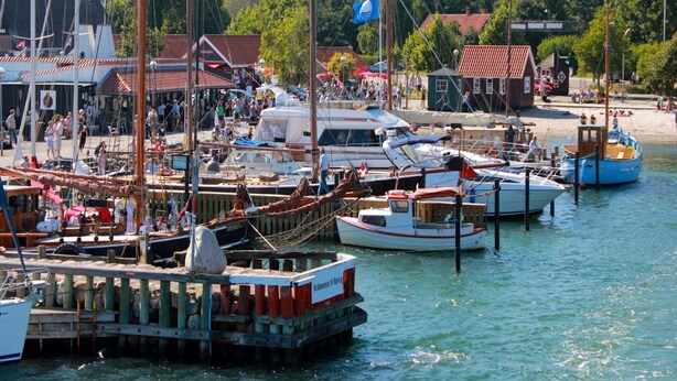 Uma marina com vários barcos atracados, um pequeno cais com uma placa e um edifício com telhado vermelho ao fundo.