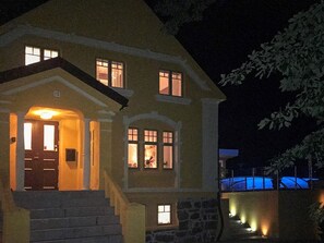 Um edifício de dois andares com fachada de pedra, entrada central com degraus e várias janelas iluminadas.