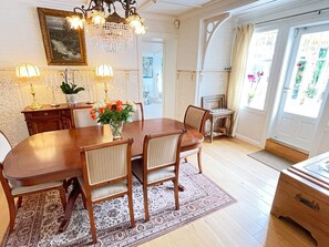 Uma sala de jantar com mesa de madeira, quatro cadeiras, um vaso com flores, um lustre e um quadro na parede.