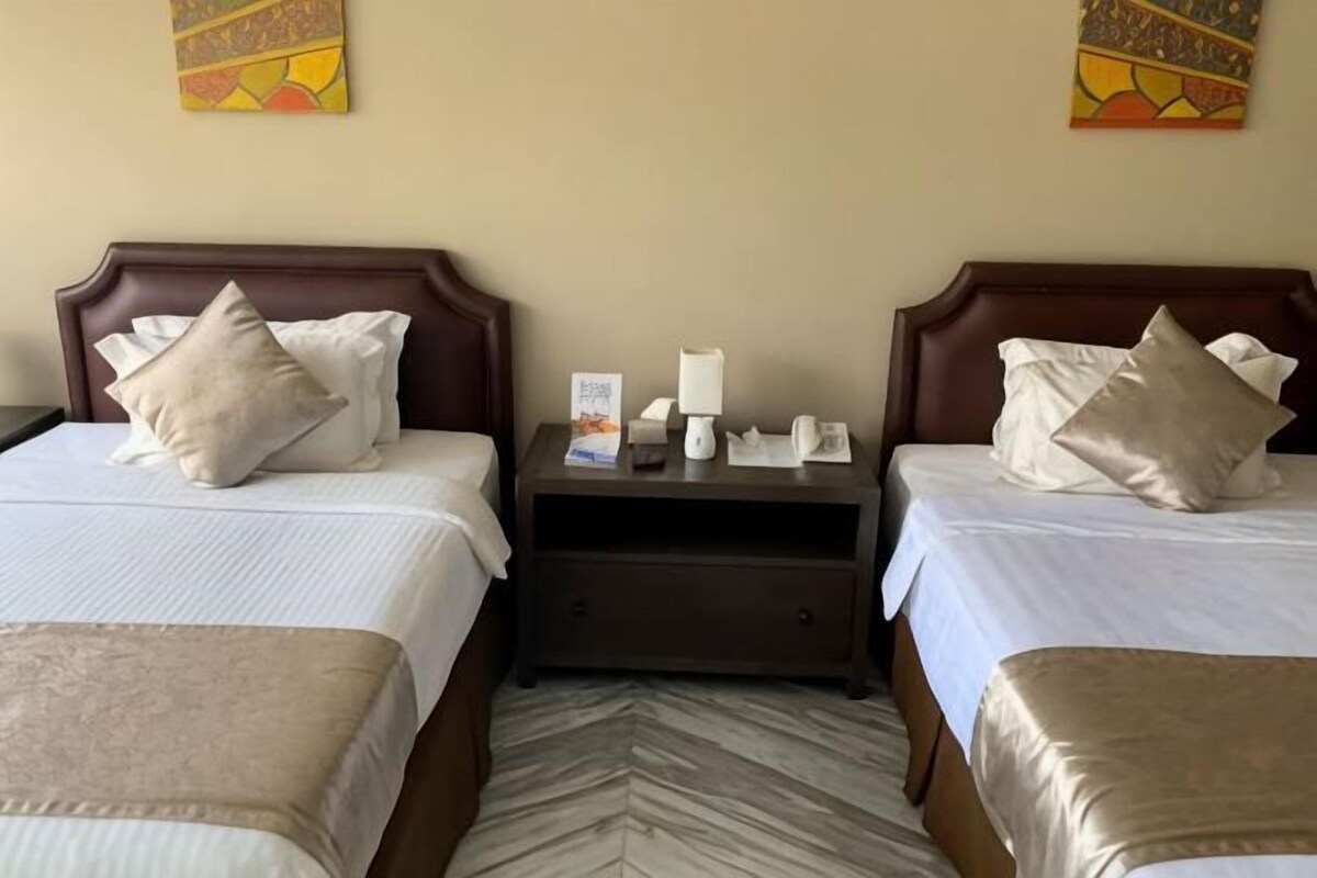 Quarto de hotel com duas camas, uma mesinha de cabeceira e piso com padrão espinha de peixe.