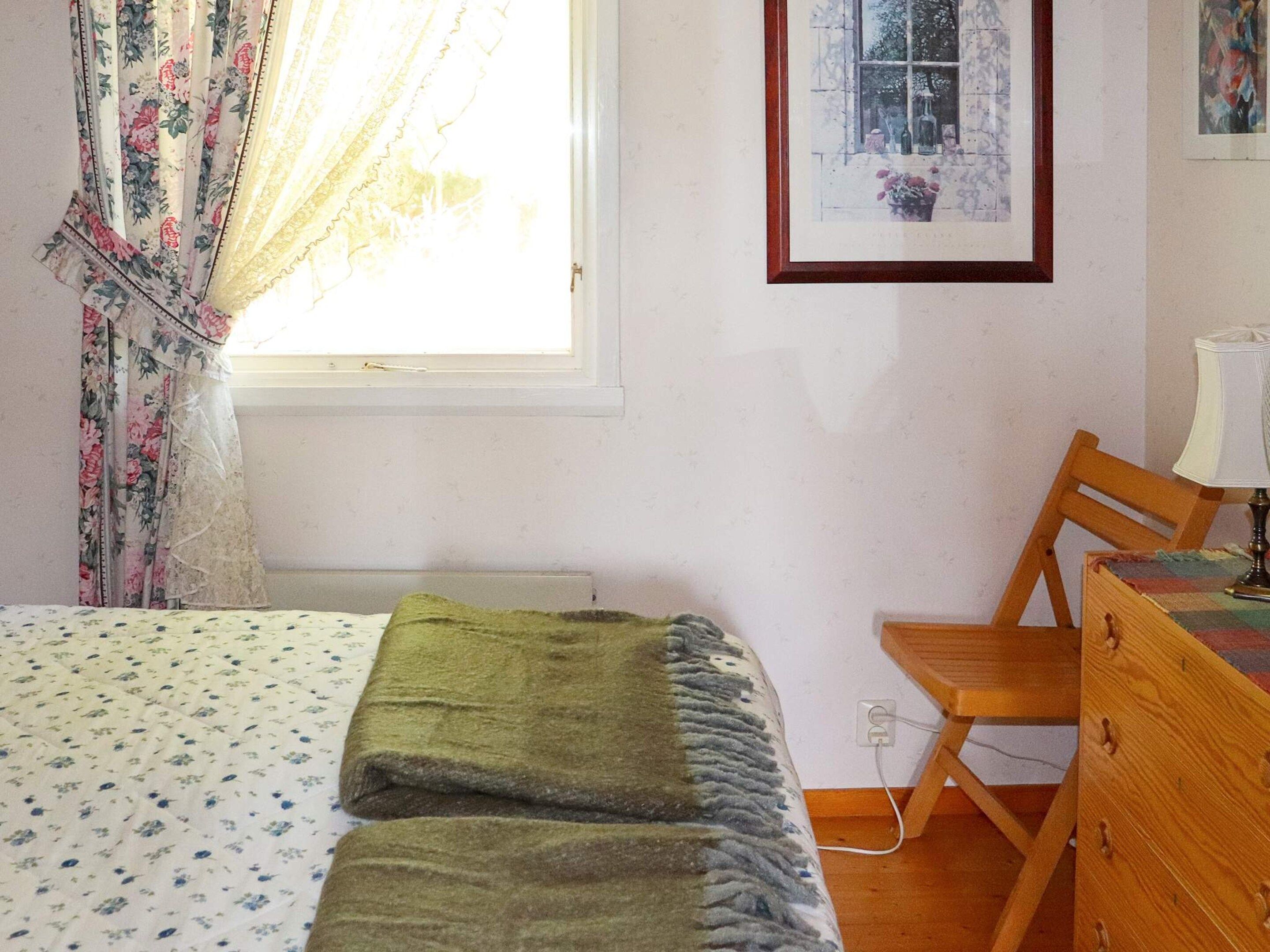 Quarto com cama, cadeira de madeira, escrivaninha com abajur, quadro na parede e janela com cortinas.
