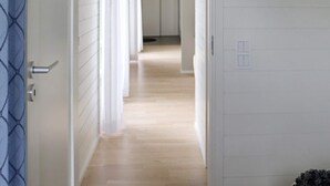 Corredor com piso de madeira e paredes brancas, conduzindo a um quarto com porta.