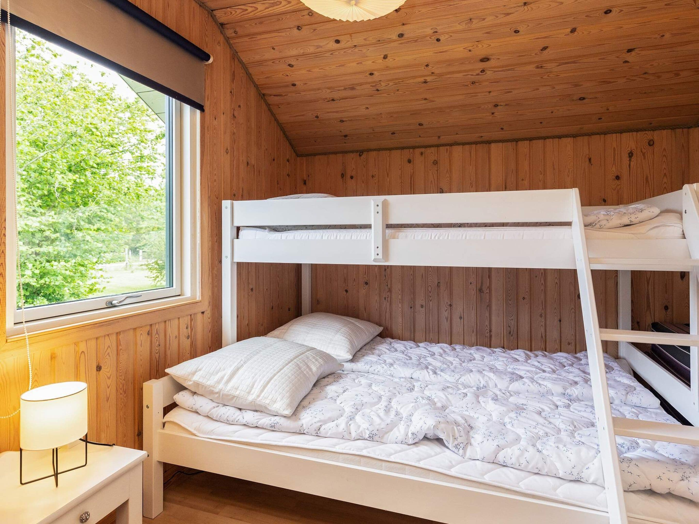 Uma cama de beliche com estrutura e roupa de cama brancas, uma mesa de cabeceira com abajur e uma janela com vista para o verde.