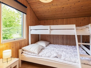 Uma cama de beliche com estrutura e roupa de cama brancas, uma mesa de cabeceira com abajur e uma janela com vista para o verde.