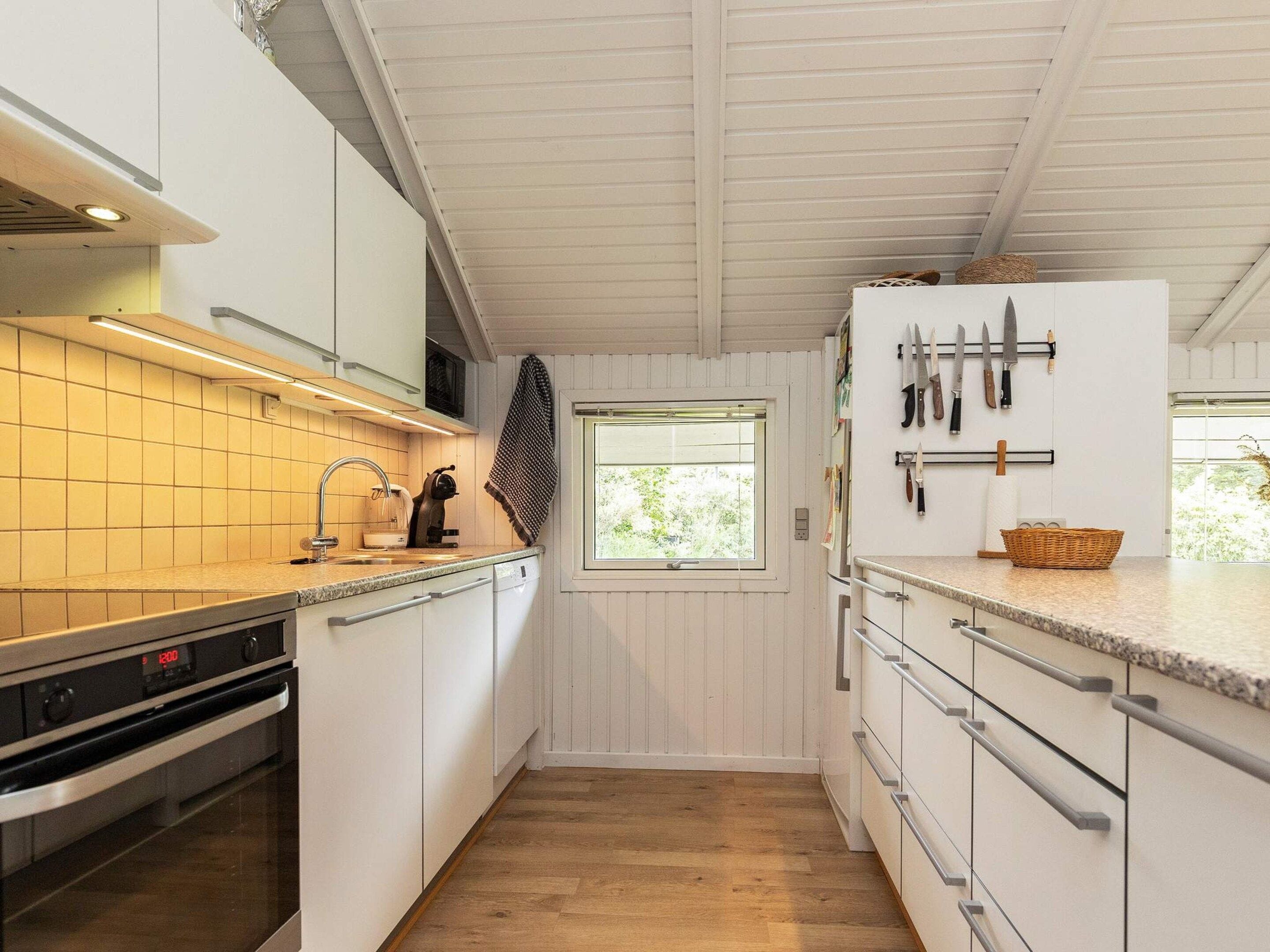 Cozinha compacta com forno embutido, micro-ondas e pia. Há uma janela que oferece luz natural e um suporte para facas na parede.