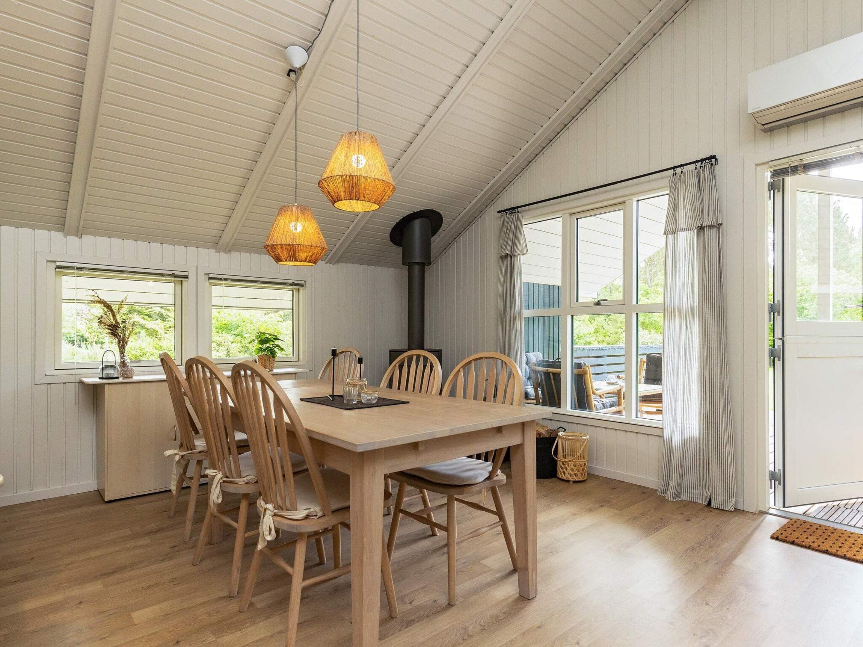 Uma área de refeições com mesa e cadeiras de madeira, luminárias pendentes, janela com cortinas e vista para o exterior.