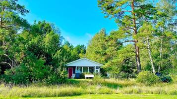 Uma cabana pequena com porta vermelha, cercada por árvores altas e um campo gramado.