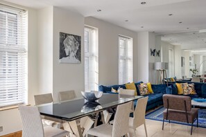 Uma sala de estar moderna com uma mesa de jantar de vidro, sofá azul e cadeiras brancas.