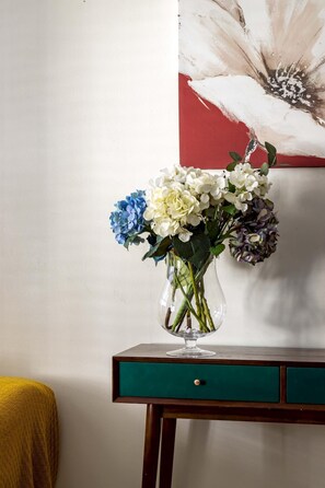 Um vaso com flores sobre uma mesa de madeira, em frente a uma parede branca com um quadro vermelho.