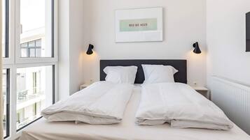 Quarto de hotel com cama, duas mesas de cabeceira, luminária na parede e uma janela.