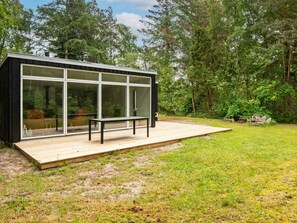 Estrutura moderna ao ar livre com deck de madeira e paredes de vidro, rodeada por uma área arborizada.