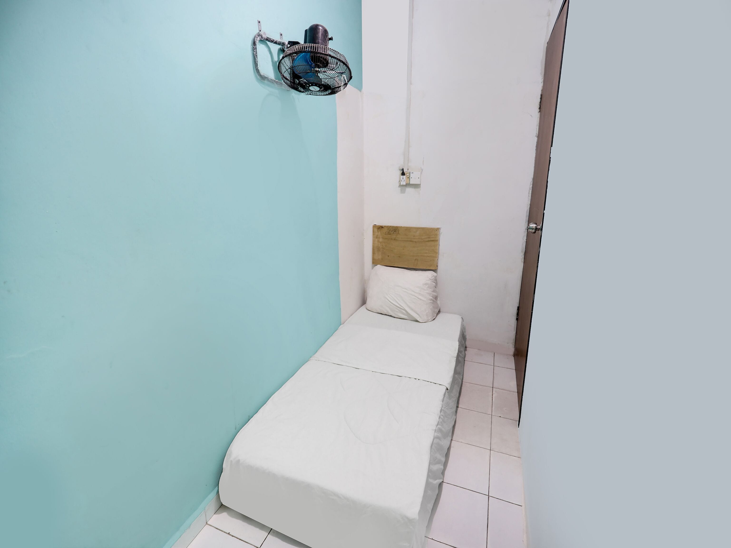 Quarto pequeno e estreito com uma cama de solteiro, um ventilador fixado na parede e uma porta.