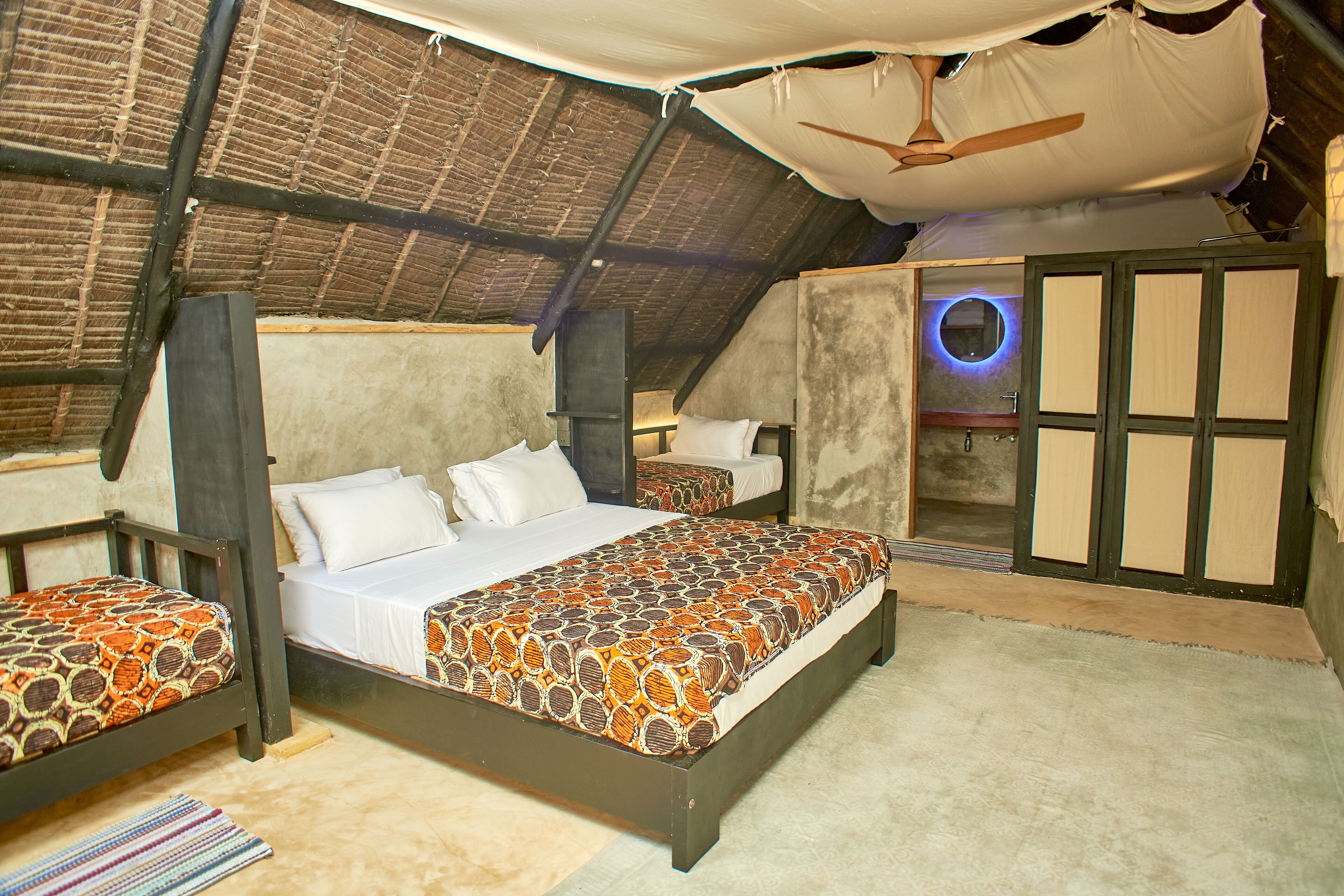 Quarto com telhado de palha, cama com roupa de cama estampada, cabeceira de madeira e uma pequena janela circular com luz azul.