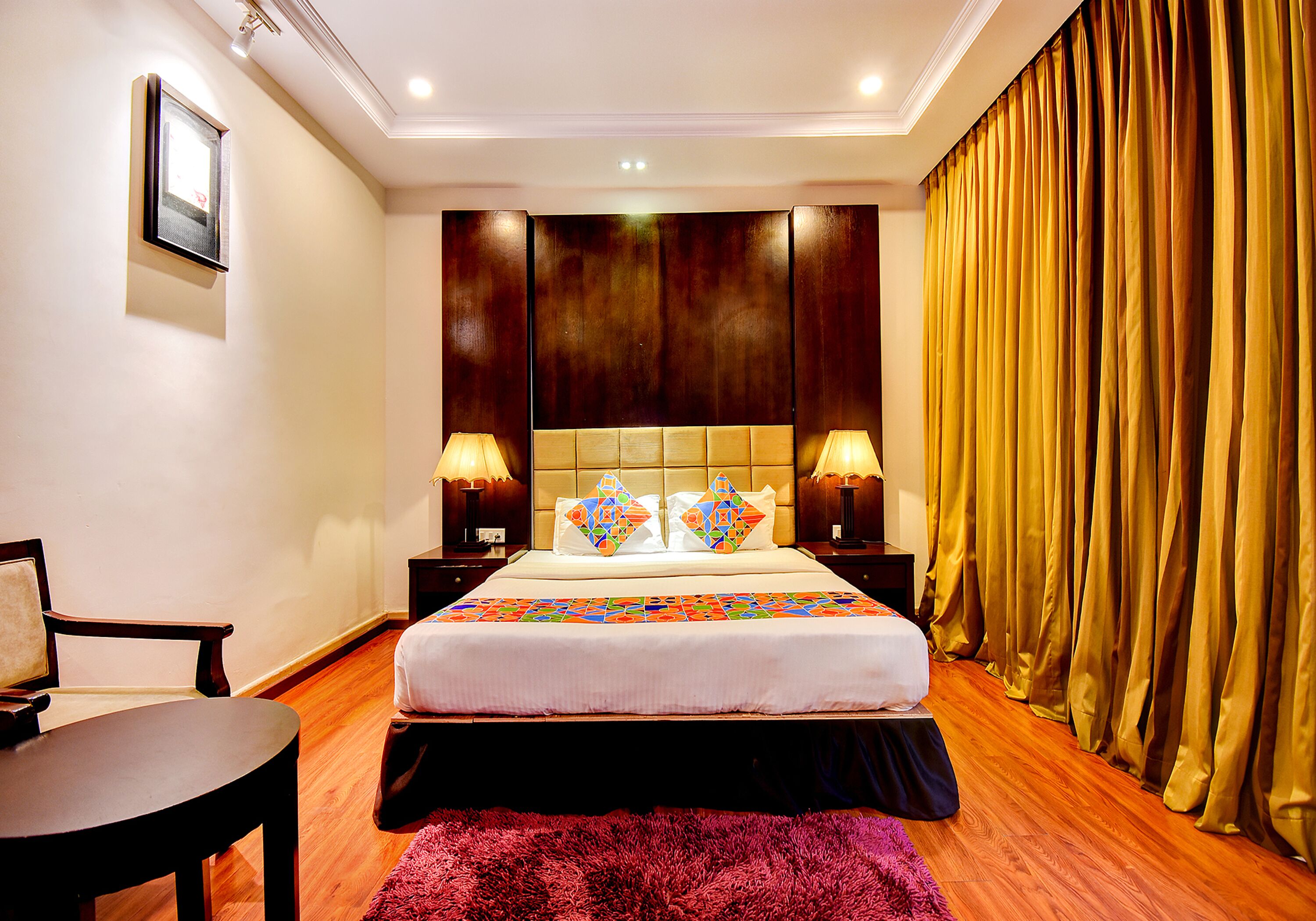 Quarto de hotel com uma cama grande, duas mesas de cabeceira com luminárias, uma cadeira de madeira e uma janela ampla com cortinas.