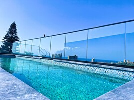 Uma piscina azul límpida com borda azulejada, guarda-corpo de vidro e vista para o oceano e prédios ao fundo.