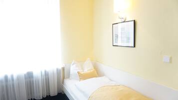 Quarto de hotel com cama, cortinas brancas, luminária fixada na parede e quadro emoldurado.
