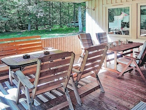Um deck de madeira ao ar livre com uma mesa, cadeiras, um banco e vista para uma área arborizada.