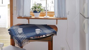 Um quarto pequeno com uma mesa, duas cadeiras, um abajur, um vaso de planta e uma janela com cortinas.