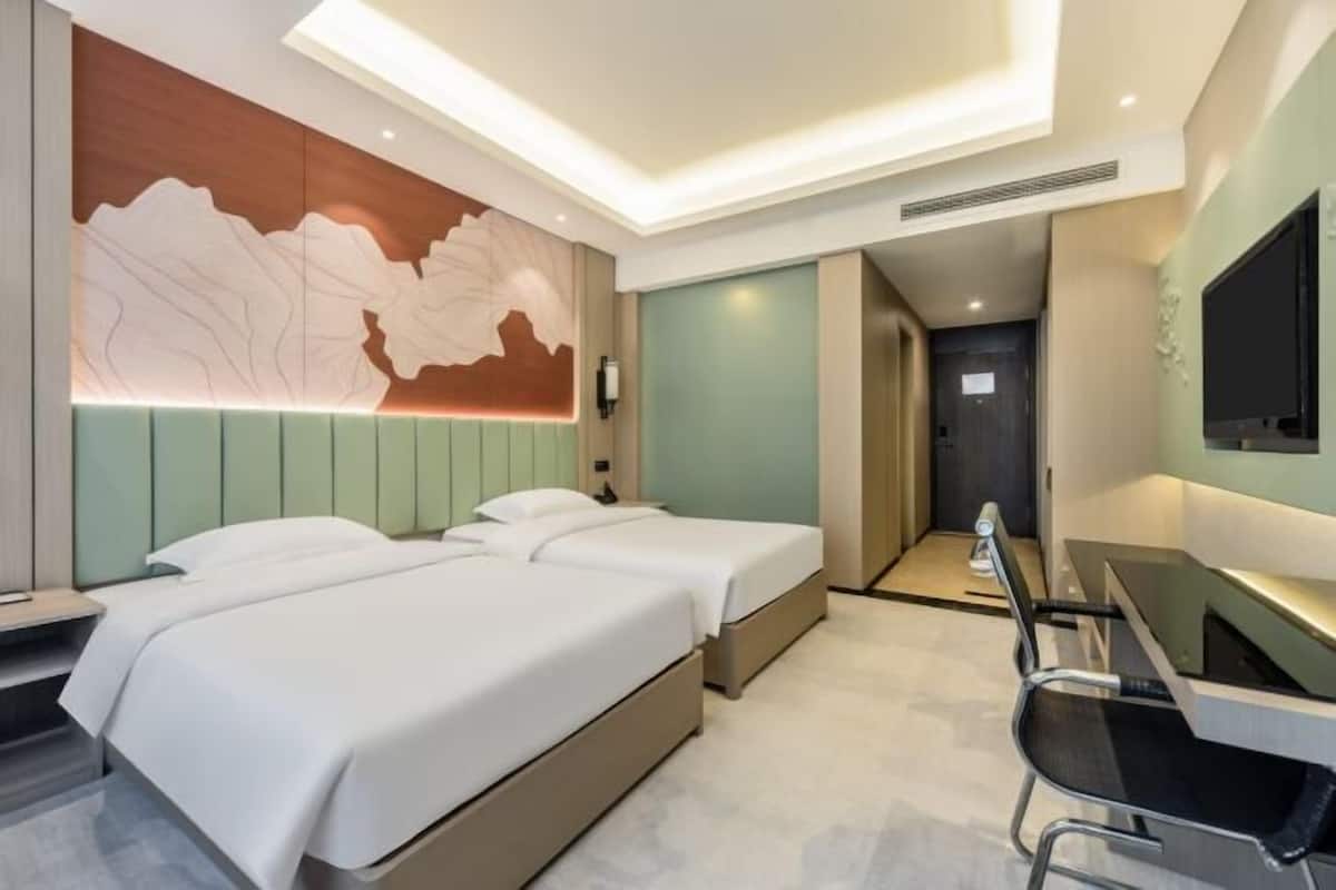 Quarto de hotel com duas camas, uma mesa, uma cadeira, uma TV e uma obra de arte na parede.