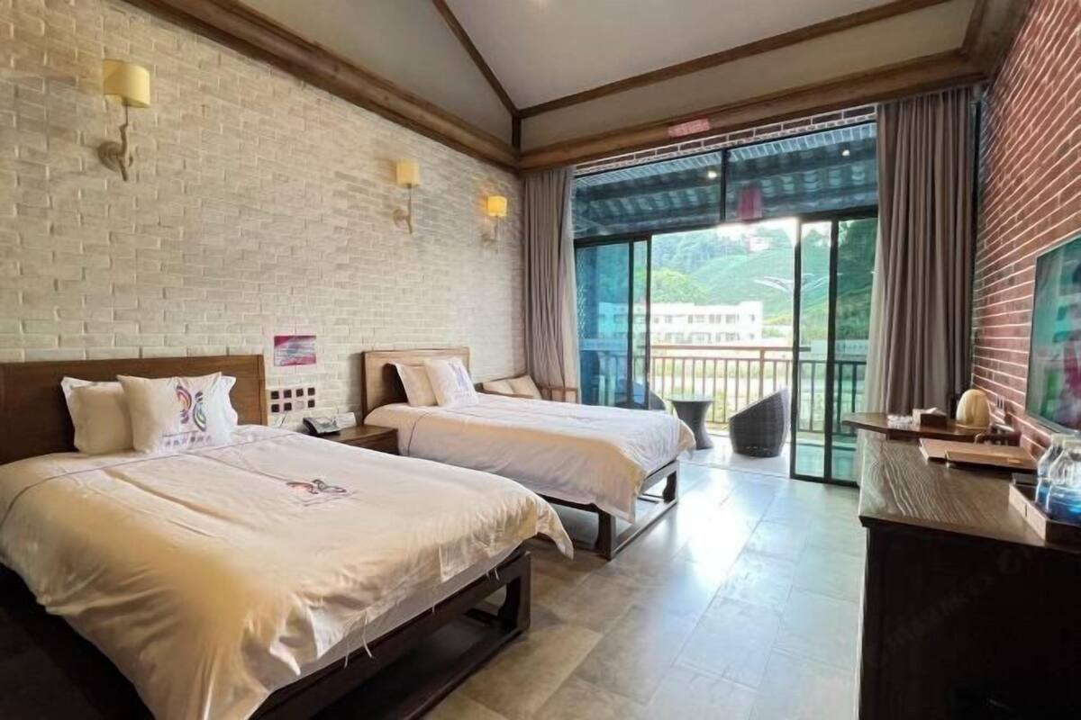 Quarto de hotel com duas camas, um painel de cabeceira de madeira, uma parede de tijolos, uma janela grande e uma varanda.