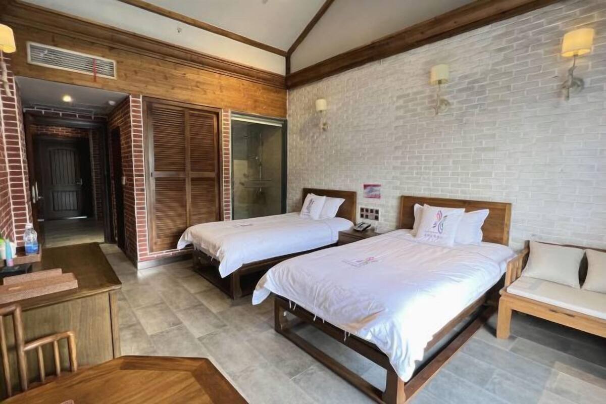 Quarto de hotel com duas camas, um banco de madeira, uma mesa pequena e uma parede de tijolos.