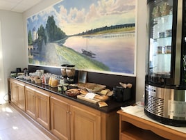 Uma bancada de cozinha com uma pintura de um lago e um barco, uma cafeteira e diversos alimentos.