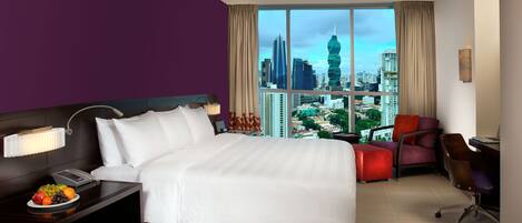 Quarto de hotel com cama, escrivaninha, cadeira e vista para a paisagem urbana.