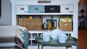 Uma bancada de cozinha com gavetas organizadas, cestos de vime, potes e uma mesa com bules.