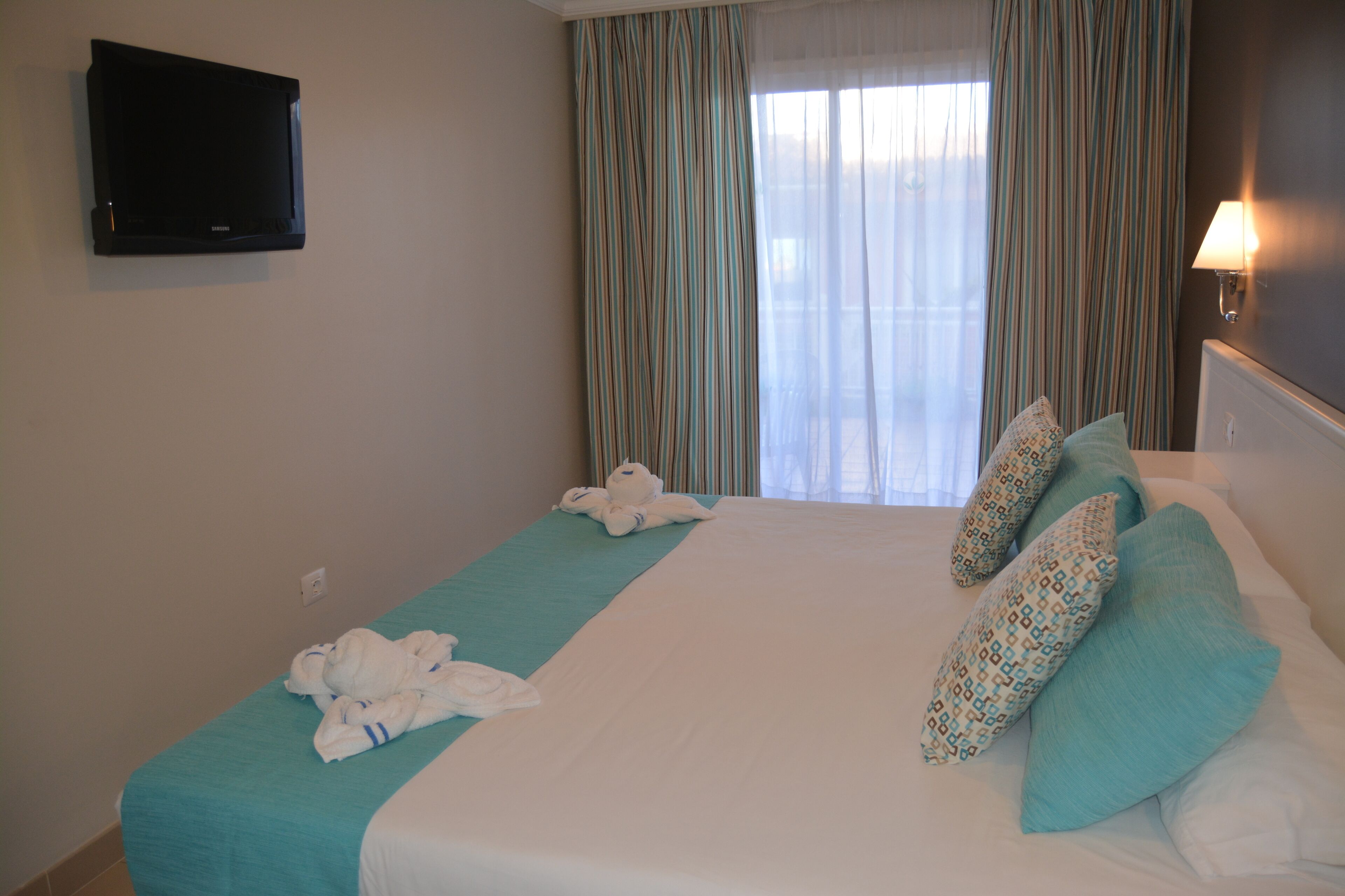 Quarto de hotel com cama, televisão de tela plana na parede e janela com cortinas.